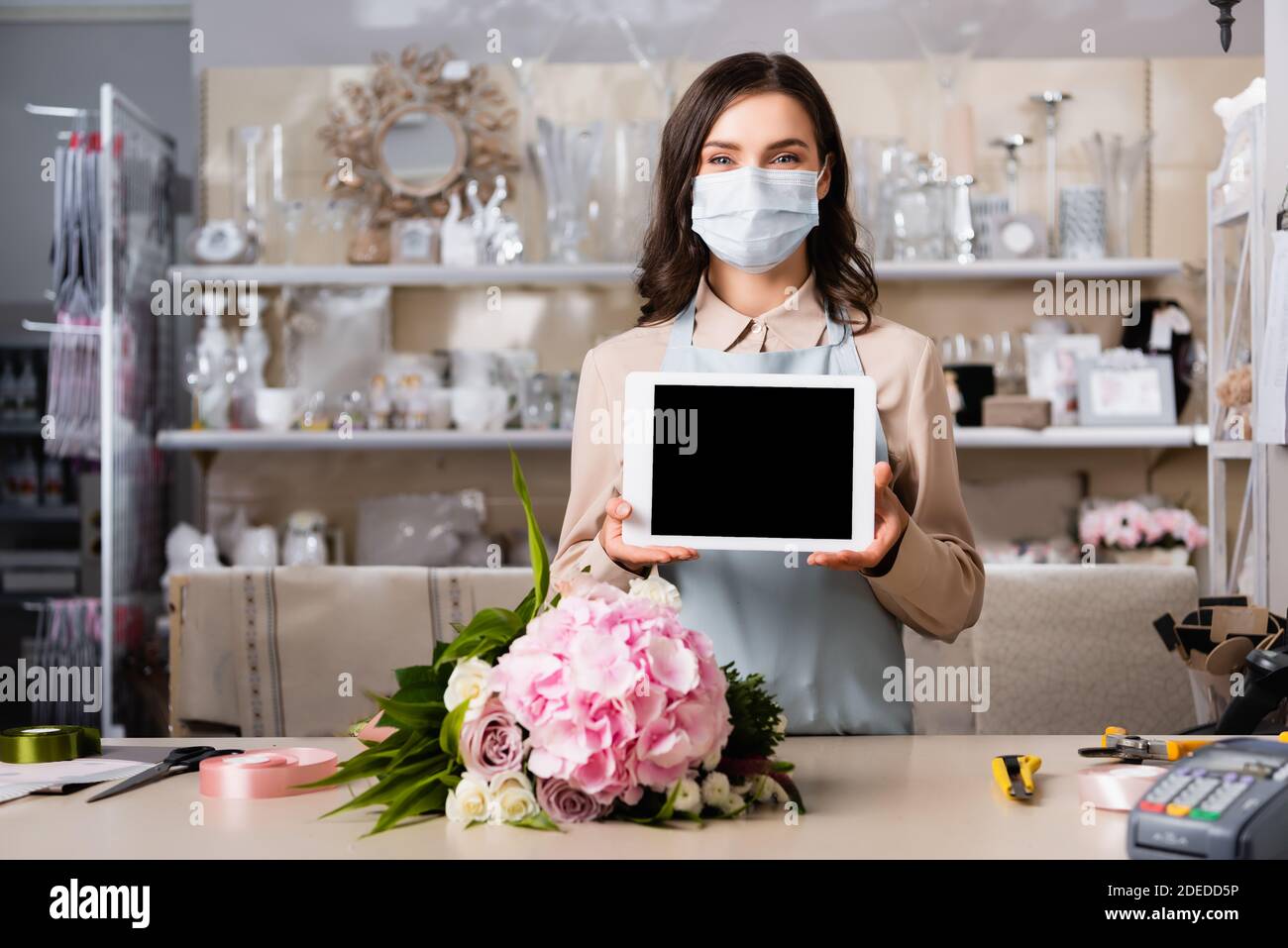 Florist in medical mask showing digital tablet near table with bouquet ...