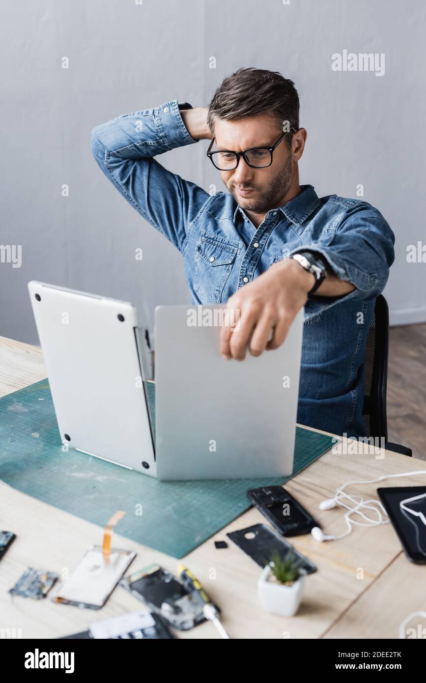 Thoughtful repairman with hand behind head looking at broken laptop at ...
