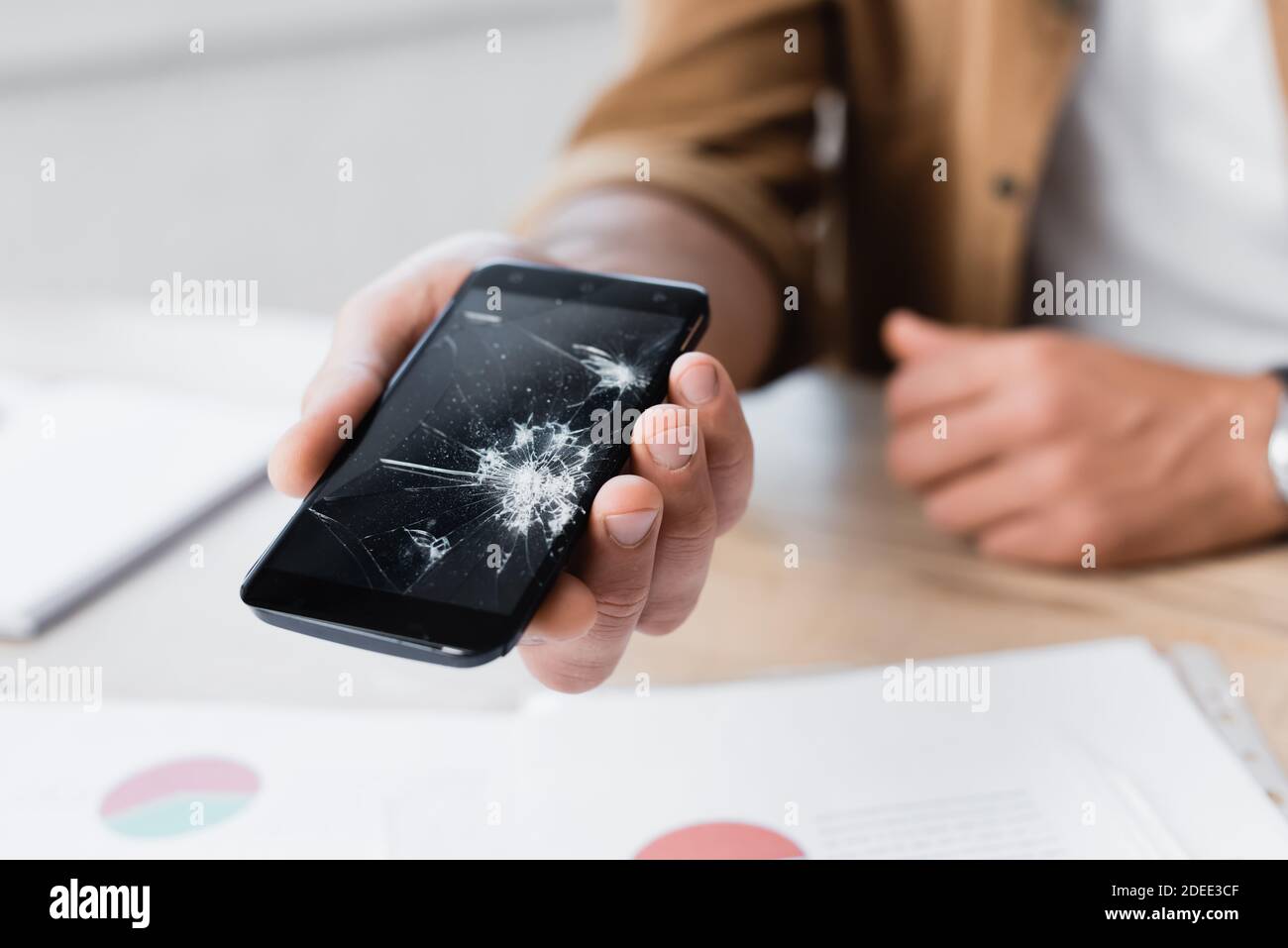 Cropped view of man holding smashed smartphone with blurred workplace ...