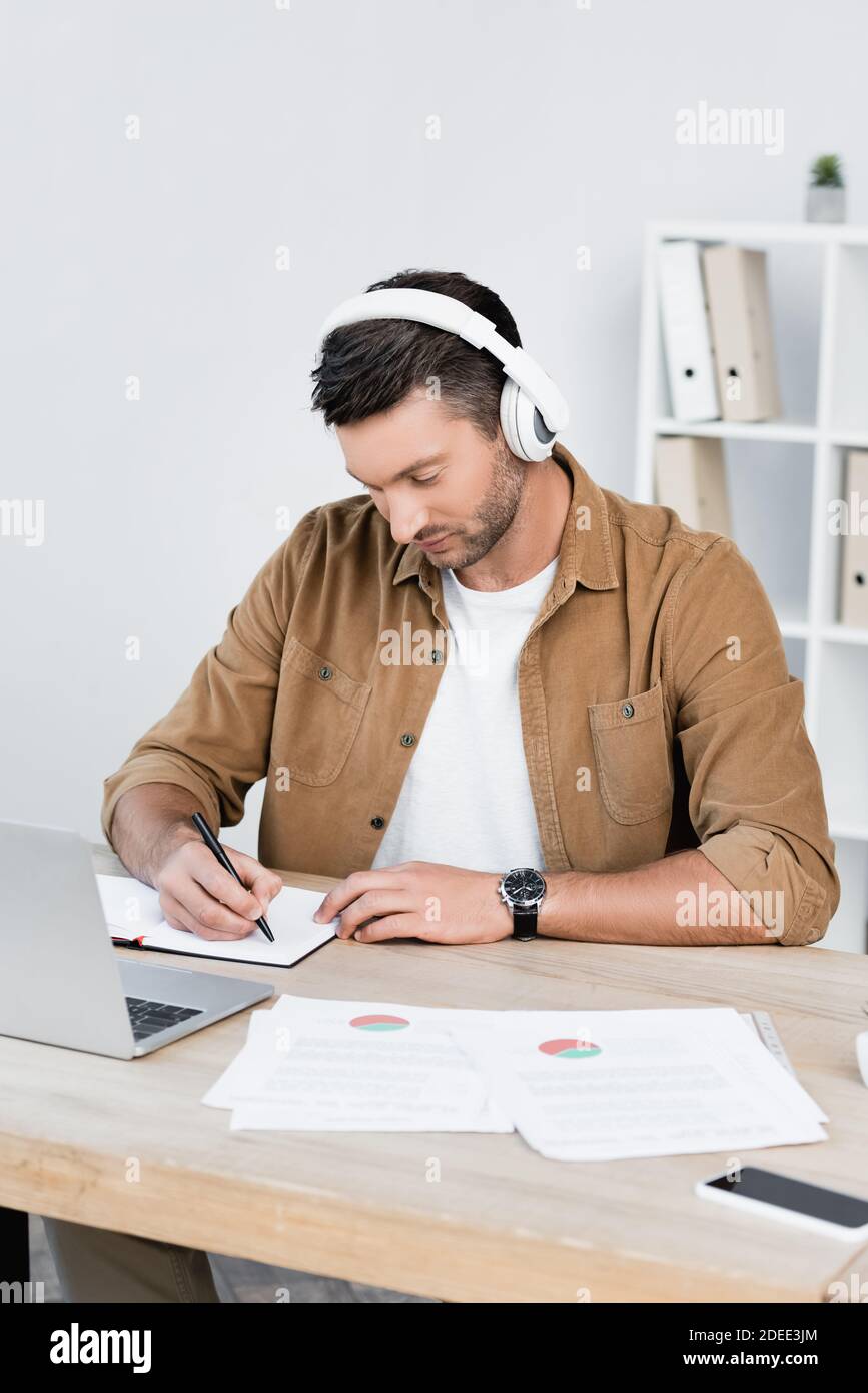 Businessman in headphones writing in notebook while sitting at ...