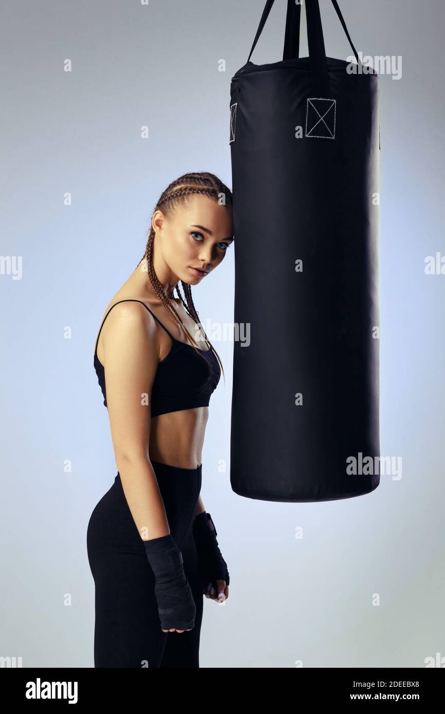 Athletic Woman In Black Boxing Bandages Leaning On Punching Bag On Gray