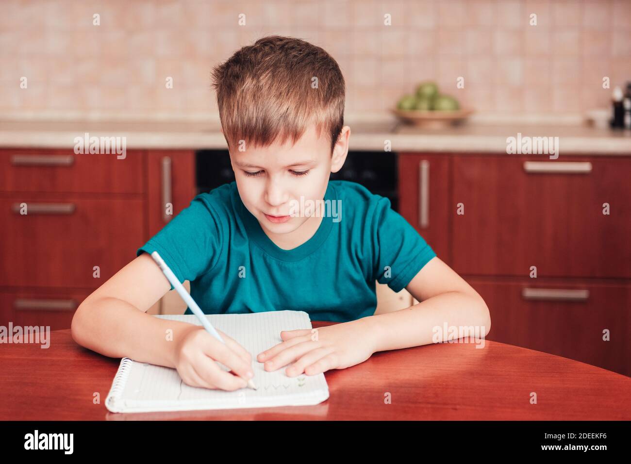 Cute child writing letters in notebook sitting by table in kitchen ...