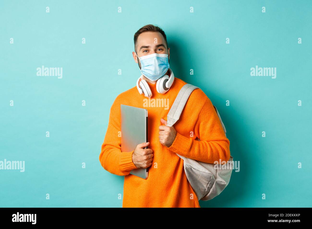 Handsome student in face mask, holding backpack and laptop, going work ...