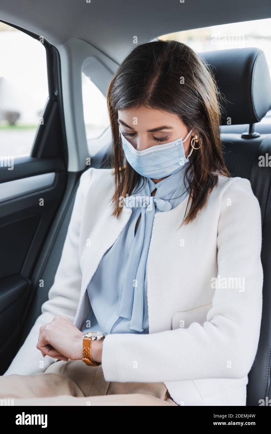 stylish woman in medical mask looking at wristwatch while riding in car ...