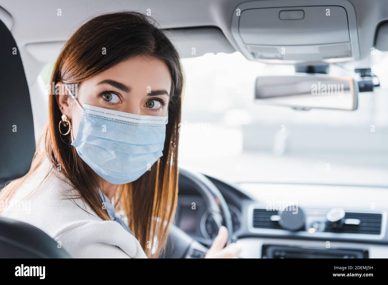 young woman in medical mask looking back while driving car on blurred ...