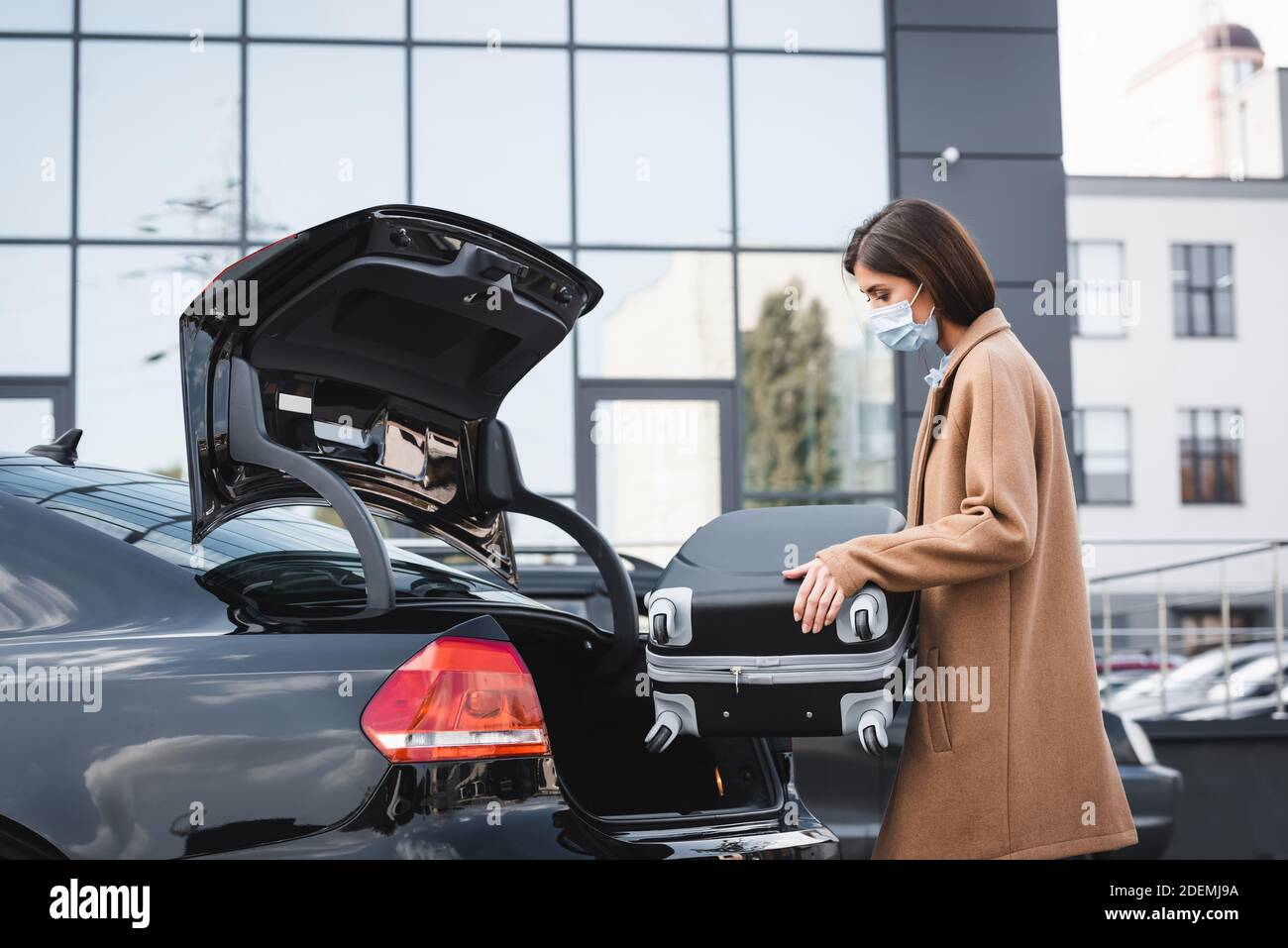 woman in trench coat and medical mask putting suitcase in car trunk ...