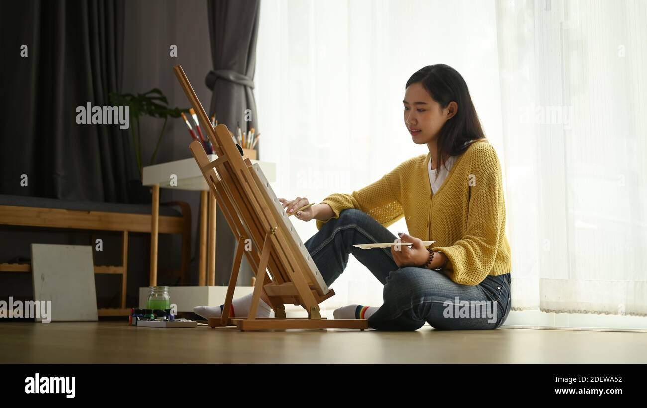 A pretty talented female painter sitting on floor and painting on easel ...
