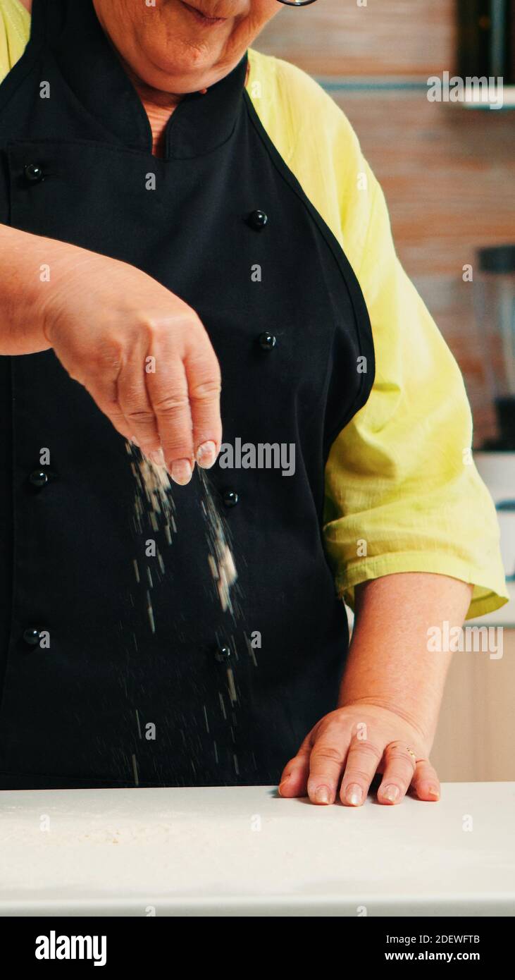 Old lady preparing food in home kitchen spreading flour for baking a ...