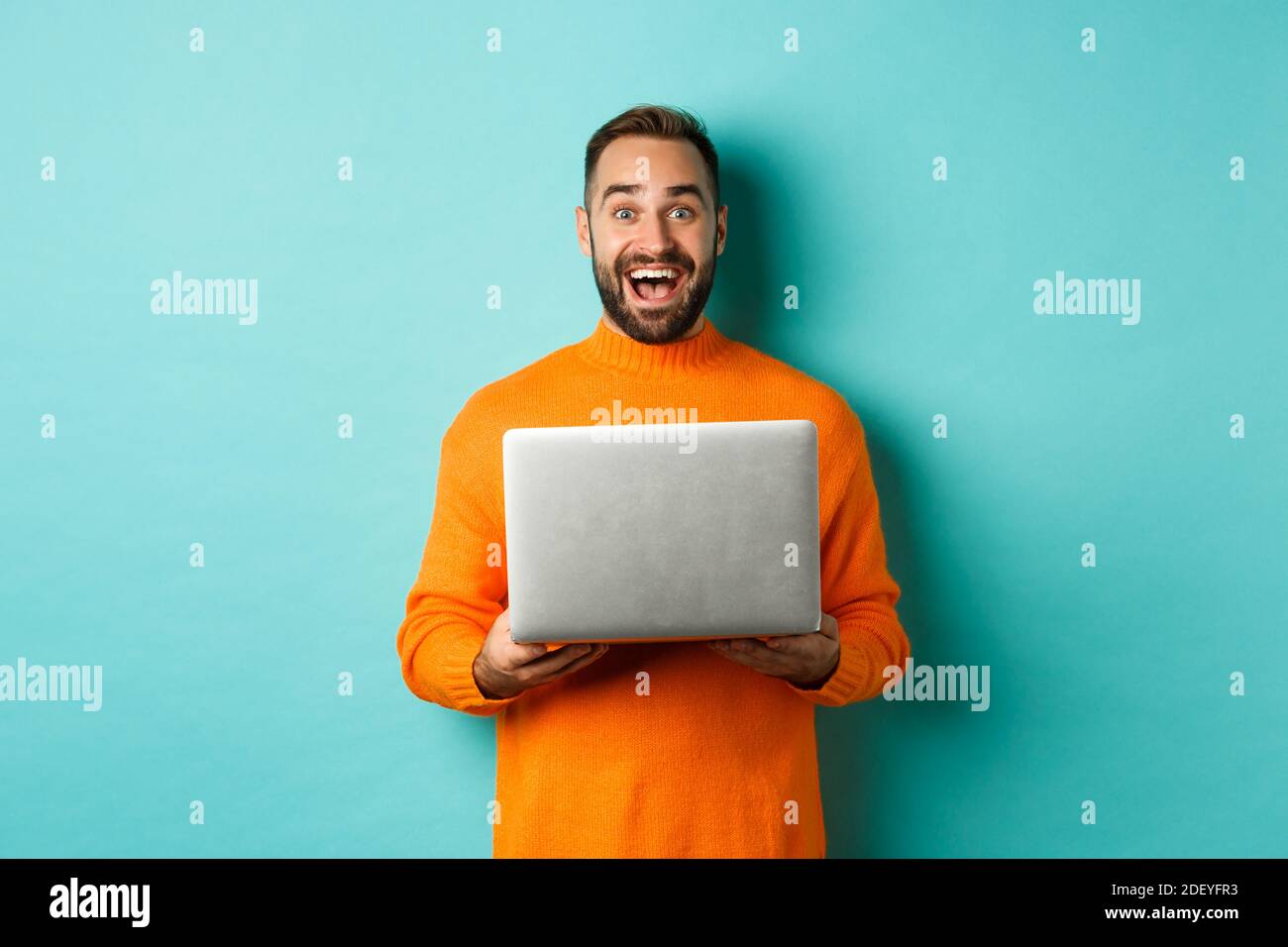 Happy man using laptop and looking excited at camera, standing with ...