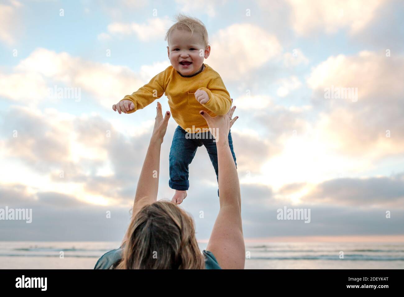 Mom tossing happy baby high into air with ocean sunset as backdrop ...