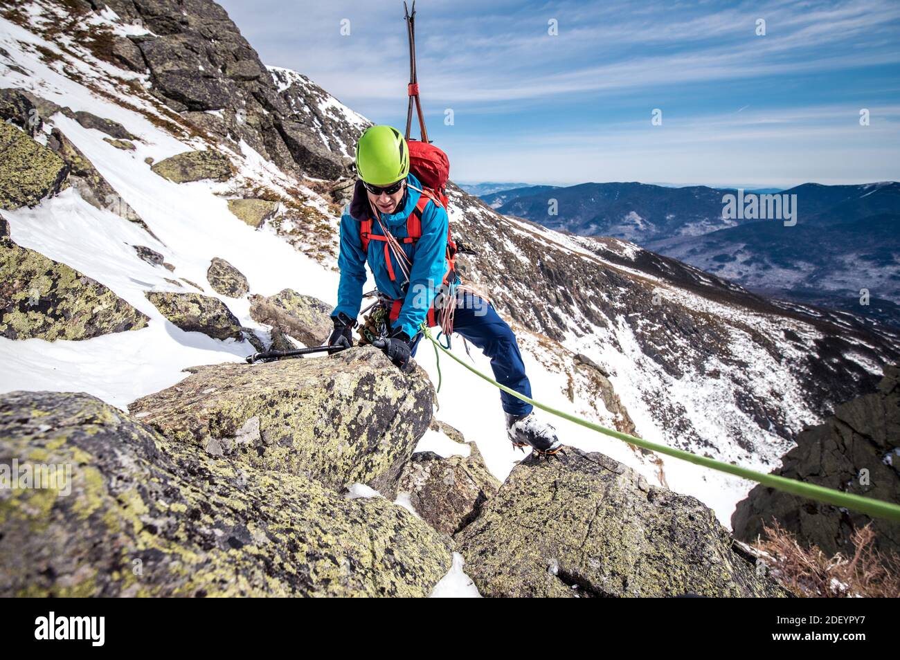 Man climbing rocks above steep snow gully with climbing rope and skis ...