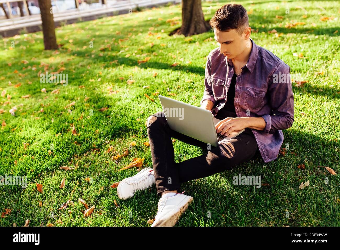 A young man in a plaid shirt, sitting on a green lawn in the park ...