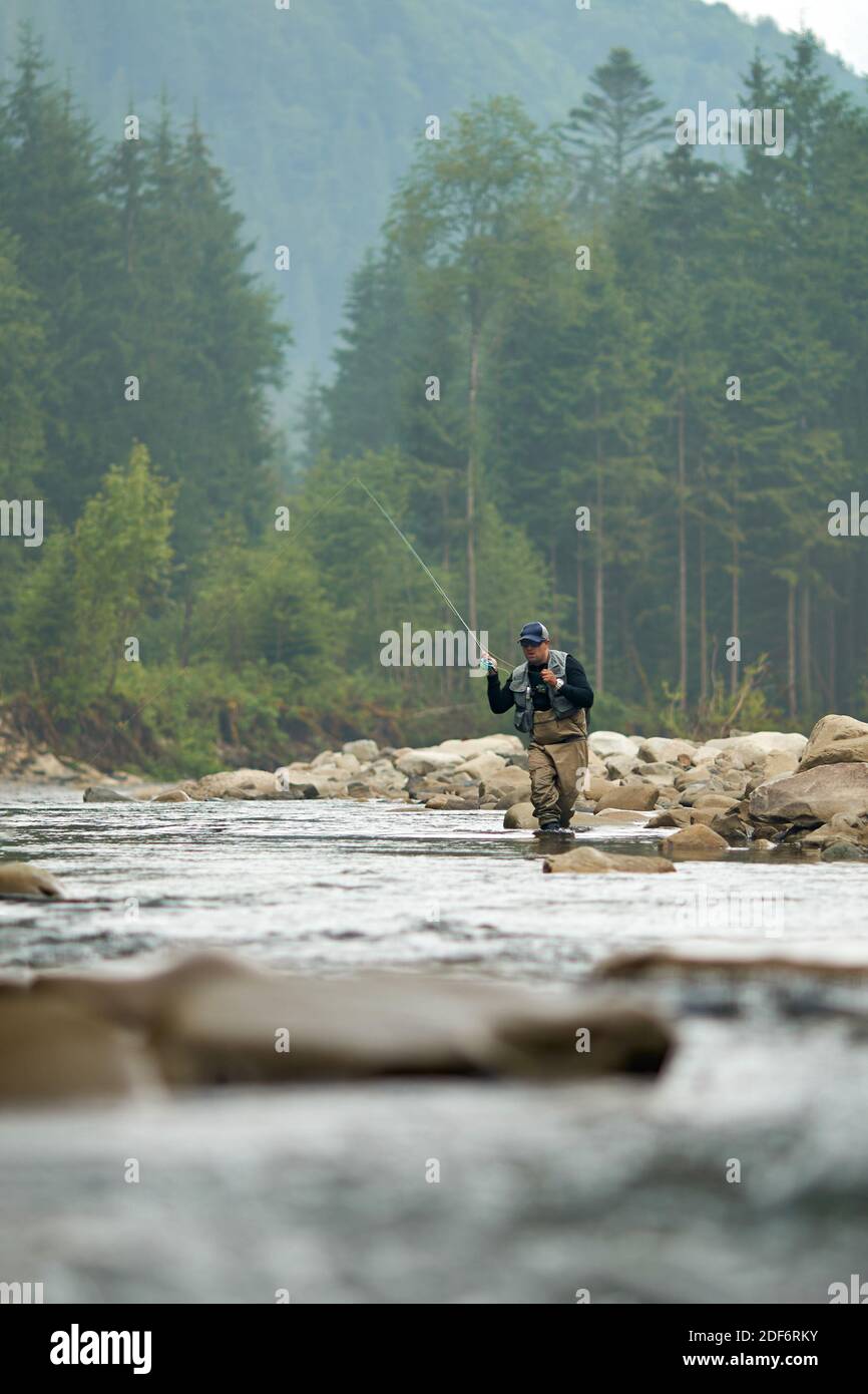 Mature man in cap, glasses and special uniform fishing with spinning ...