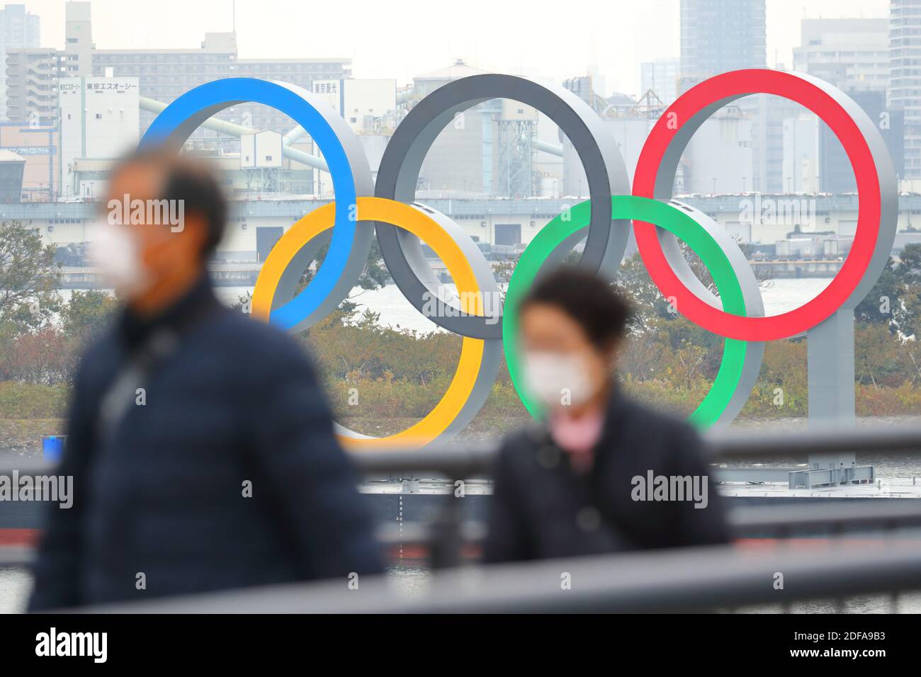 People wearing face masks walk in front of the giant Olympic rings ...
