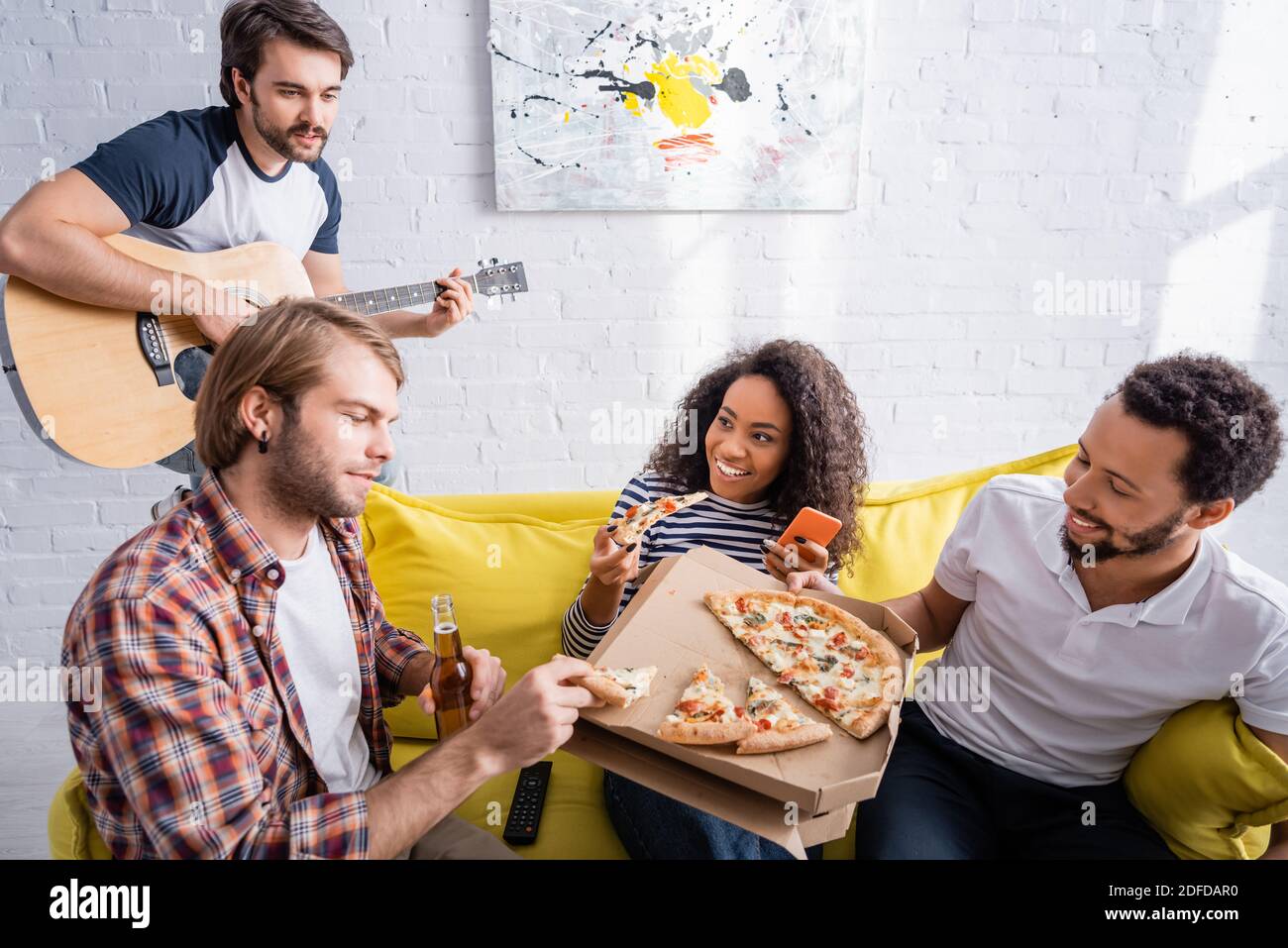 multicultural friends eating pizza on sofa while young man playing ...