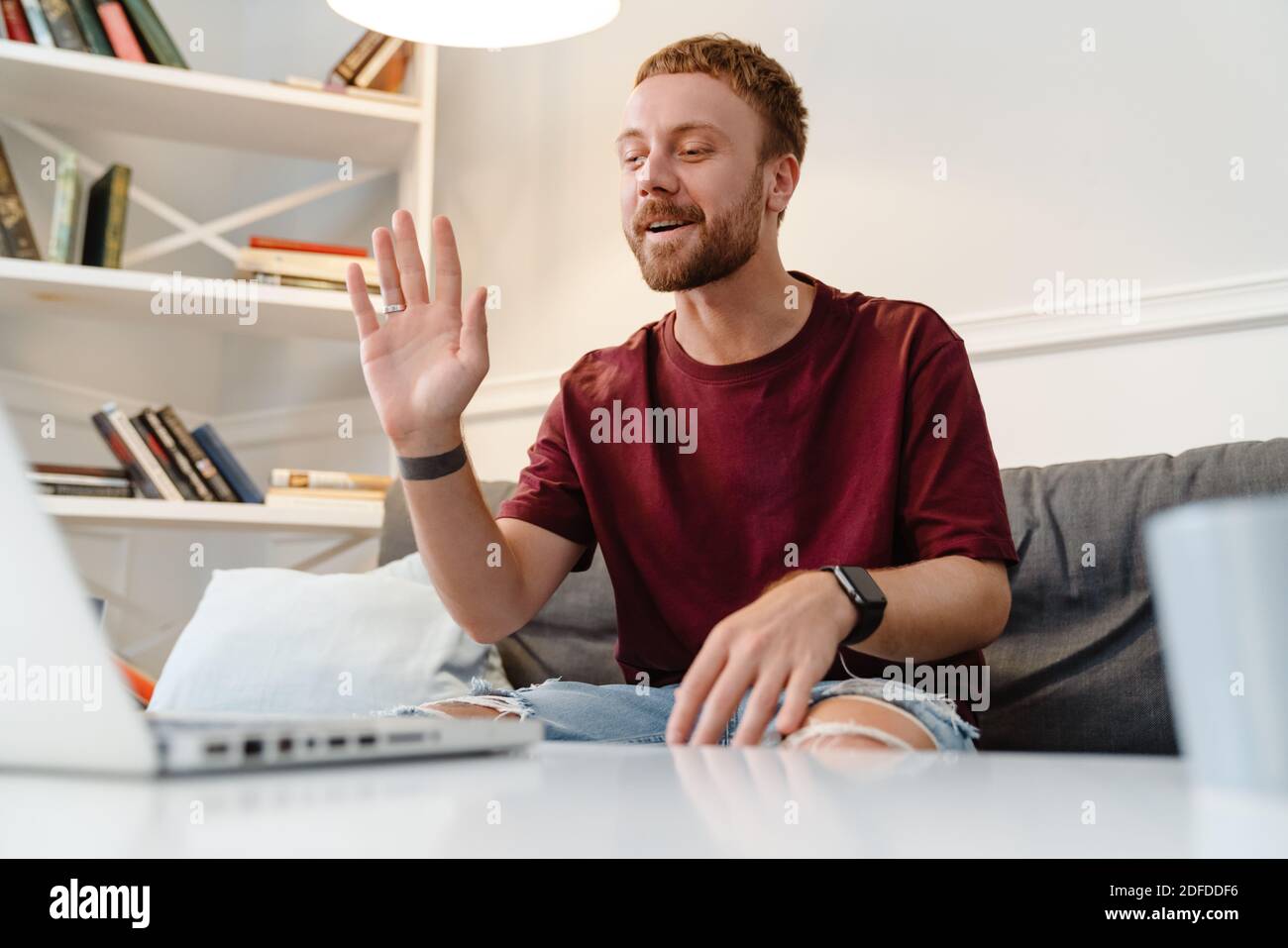Image of ginger joyful guy waving hand and using laptop while sitting ...