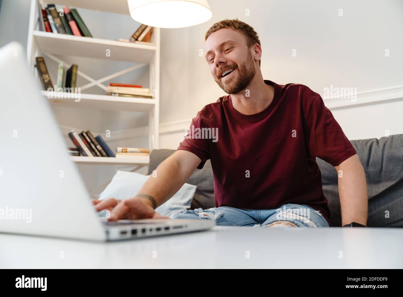Image of ginger joyful guy laughing and using laptop while sitting on ...