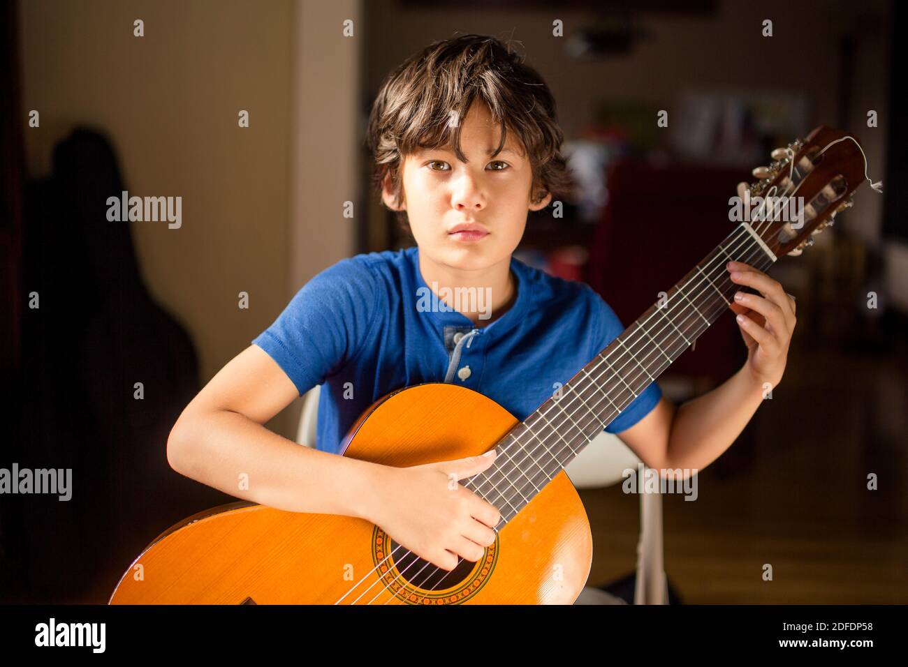 A beautiful boy with intense gaze sits in window light holding guitar ...
