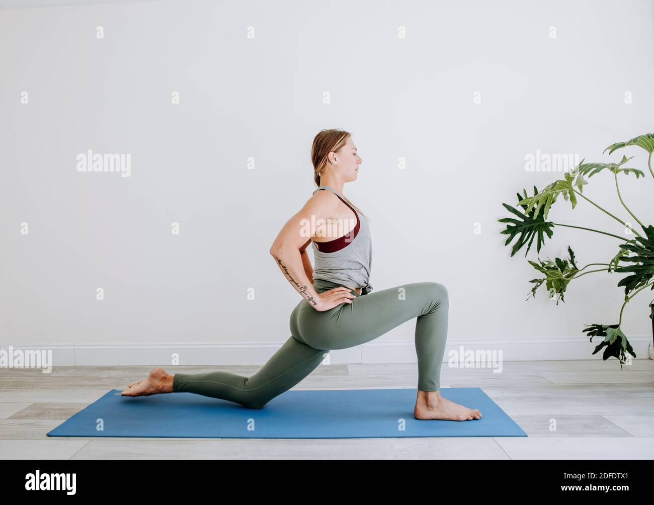 Fit white barefoot female does yoga lunge on blue mat in white room ...