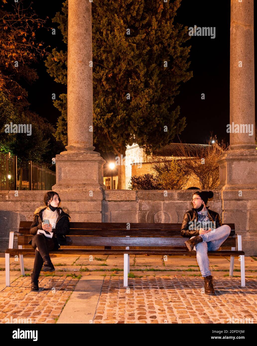 A view of two Caucasian people sitting on a bench while keeping a safe ...