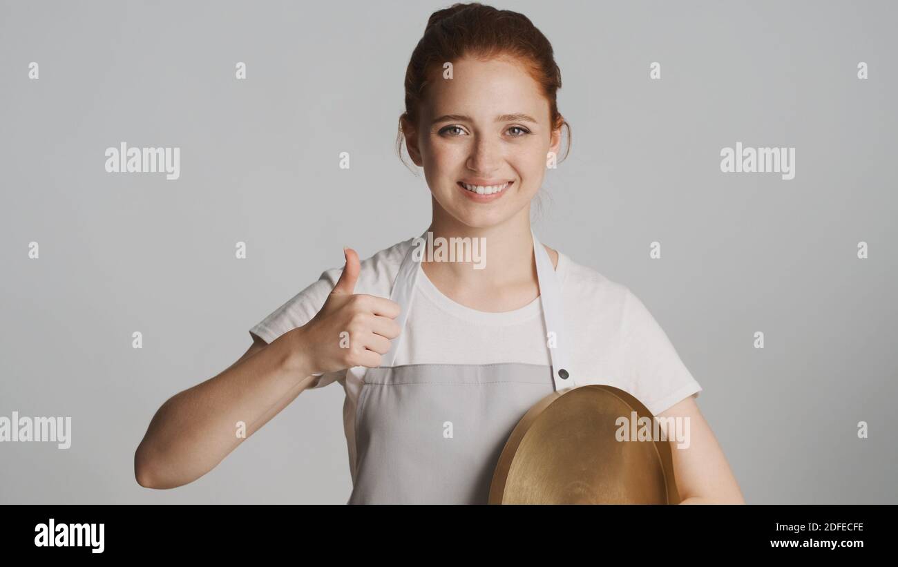 Pretty waitress in apron with tray happily showing thumb up on camera ...