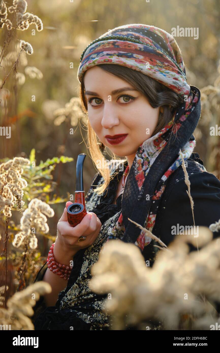 young gypsy woman smoking pipe in autumn dry grass, looking at camera ...