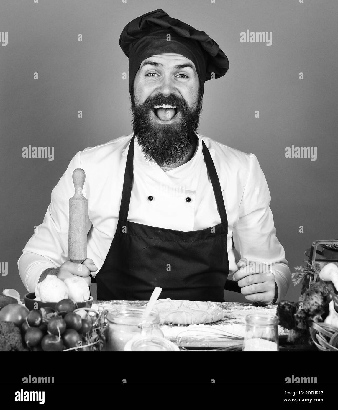 Cook with cheerful face in burgundy uniform holds rolling pin. Chef ...