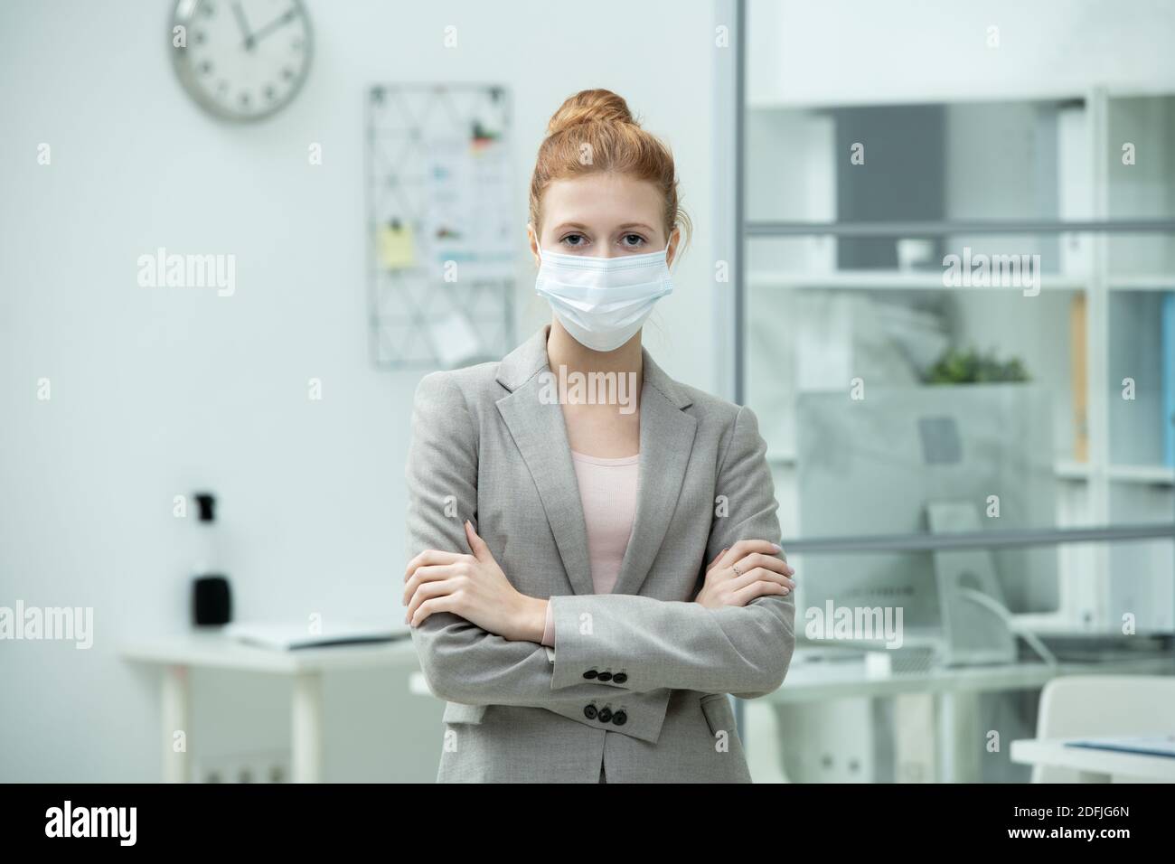 Young pretty female manager in suit and protective mask crossing arms ...