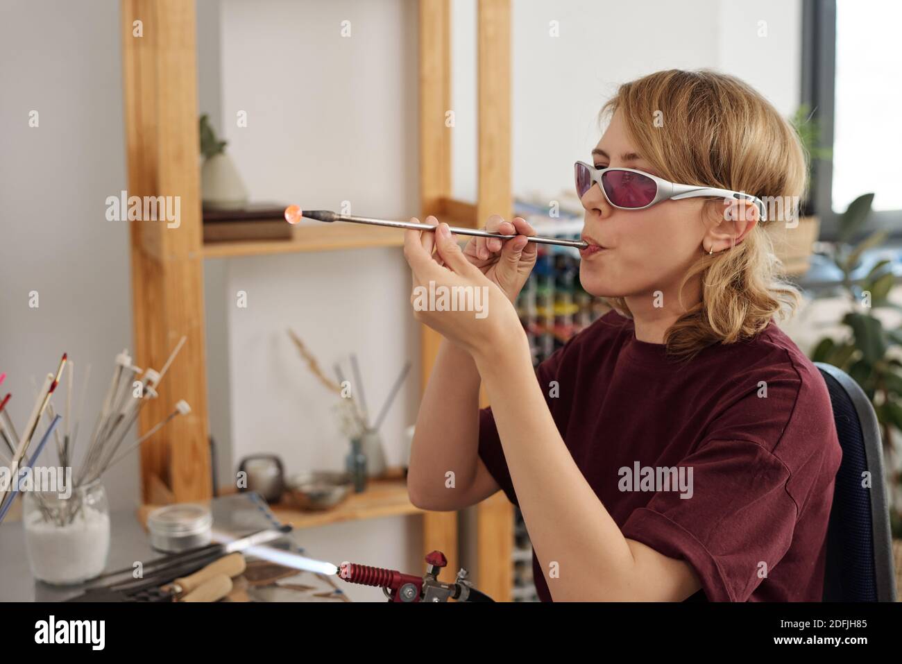 Young skilled female lampworking master blowing into long tube while ...