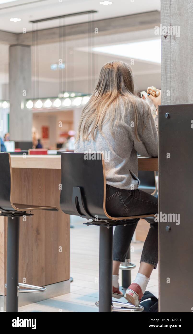 Back view of young girl sitting in indoors food court and eating an ...