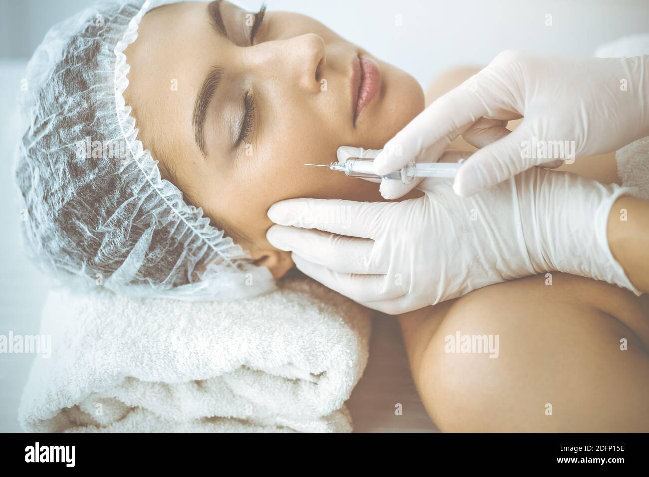 Beautician doing beauty procedure with syringe to face of young brunette woman in sunny clinic ...