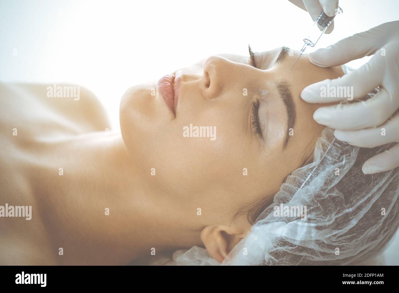 Beautician doing beauty procedure with syringe to face of young brunette woman in sunny clinic ...