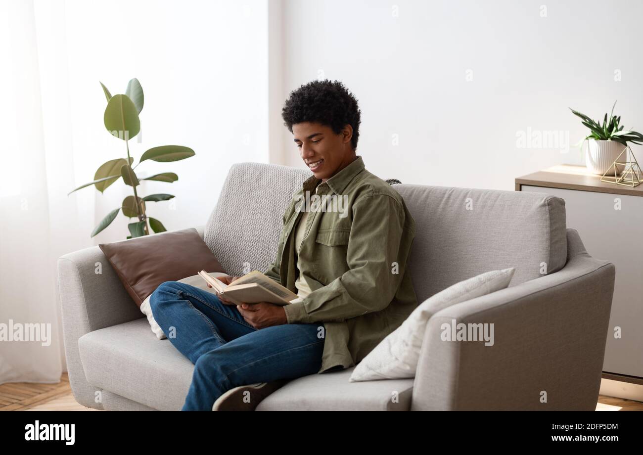 Cheerful black male student sitting on couch with book, reading and ...