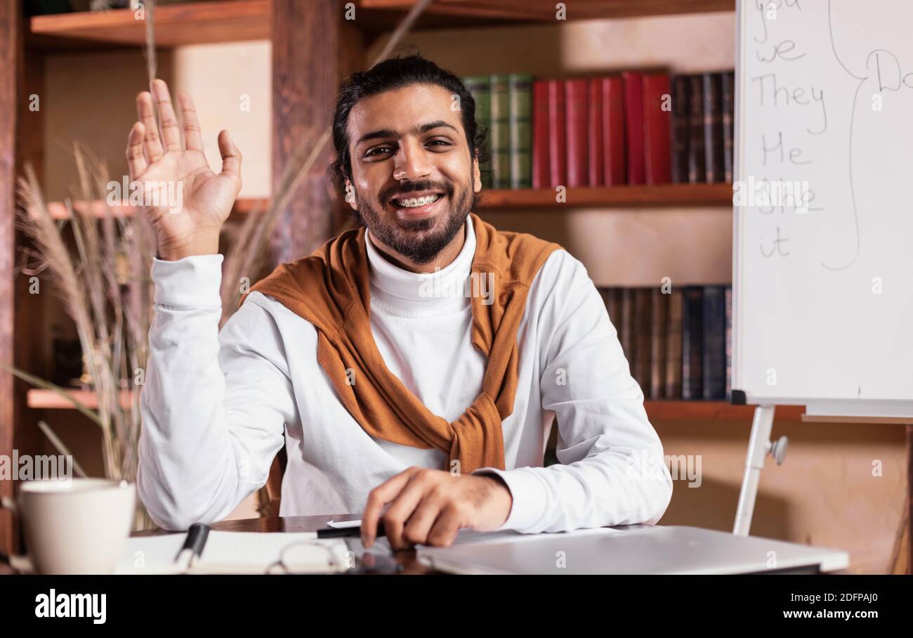 Joyful Indian Teacher Man Waving Hello Posing Sitting In Classroom ...