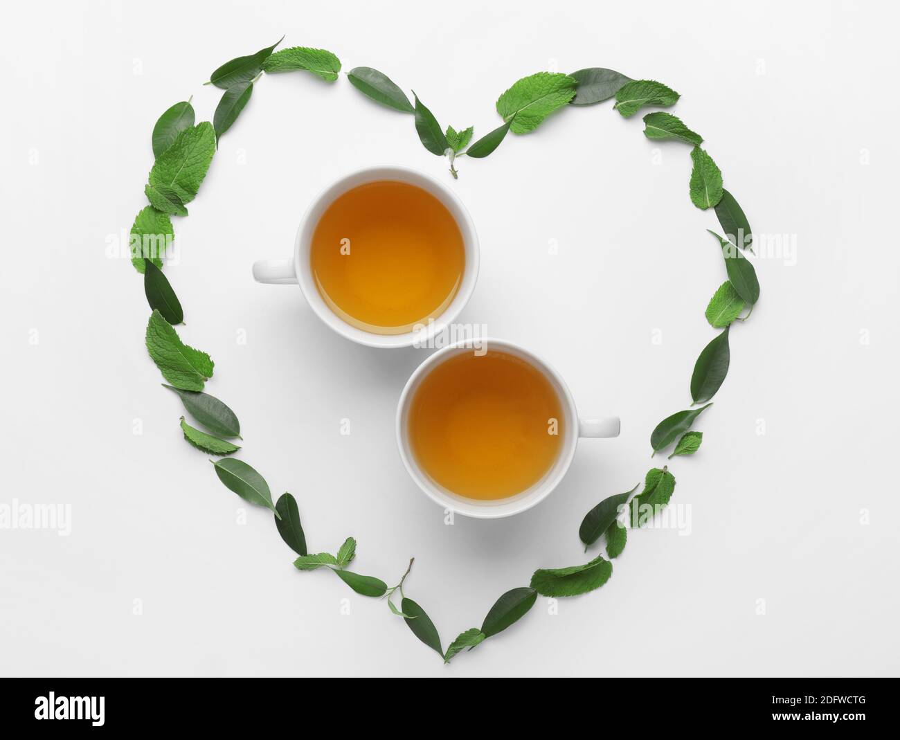Two cups of tea and heart made of green tea leaves on white background ...
