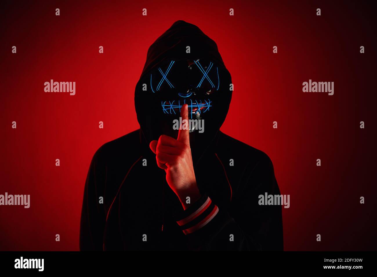Anonymous man in a hoodie hiding face behind neon glow scary mask on ...