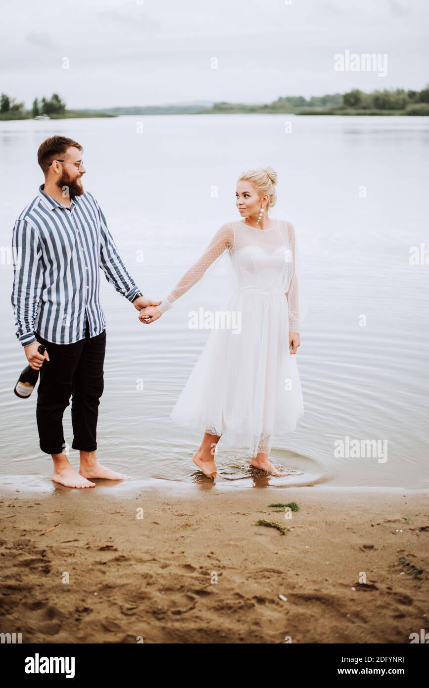 The bride and groom stroll barefoot along the river and hold hands ...