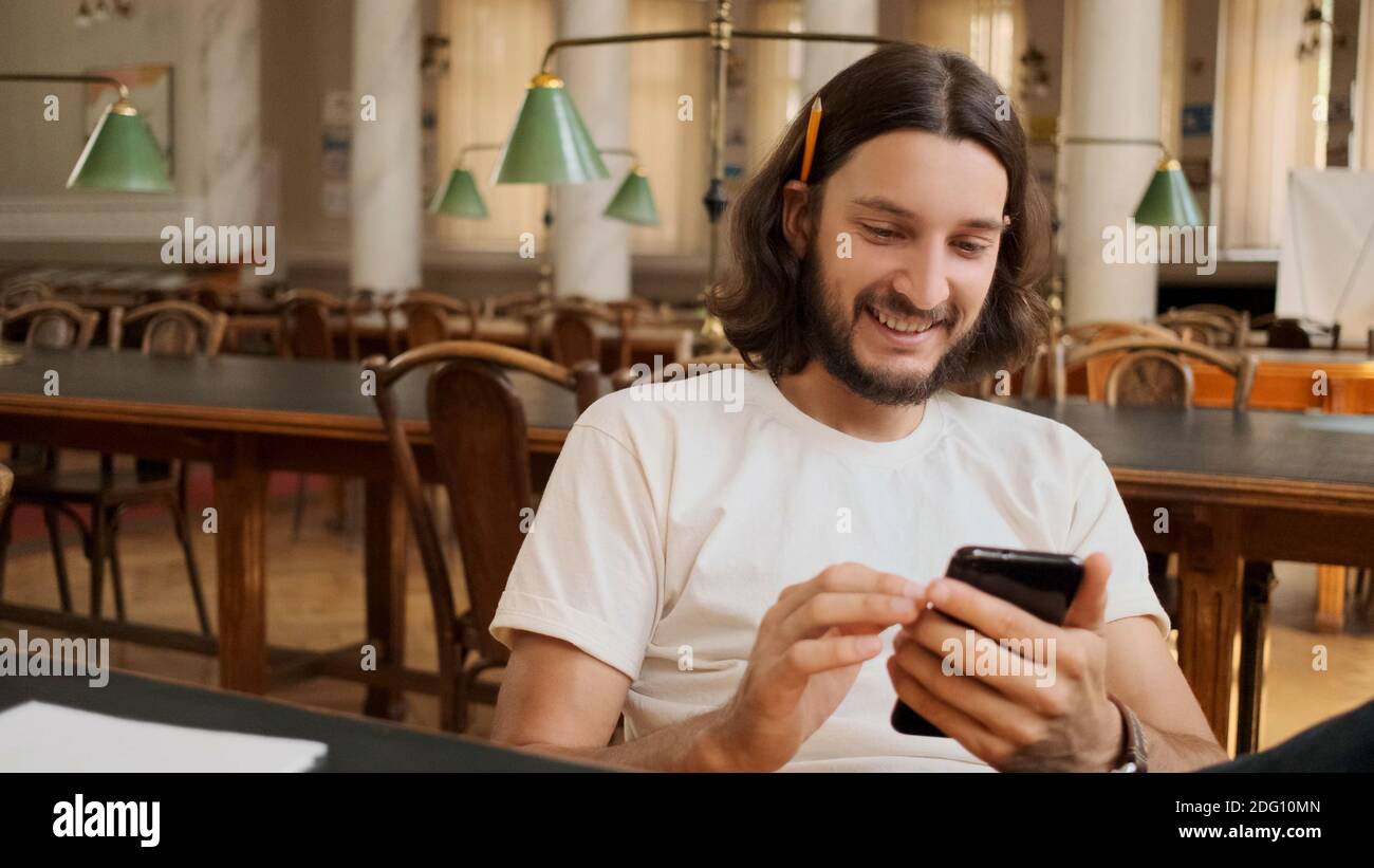 Portrait of joyful student happily using smartphone in library ...