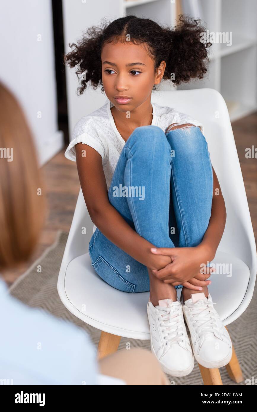 Serious african american girl hugging legs while sitting on chair ...