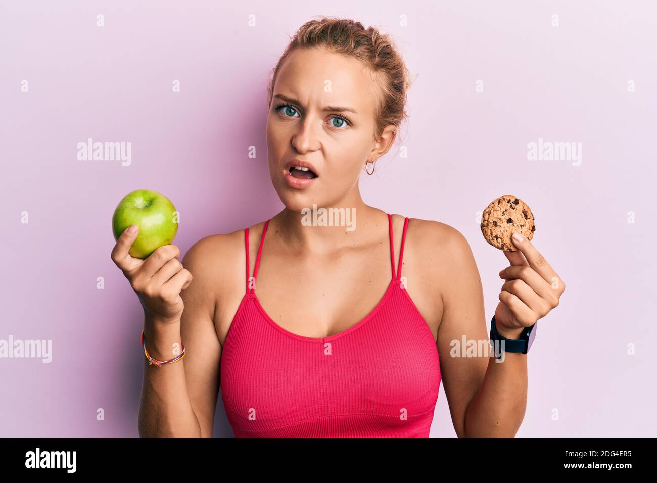 Beautiful caucasian woman holding green apple and cookie in shock face ...