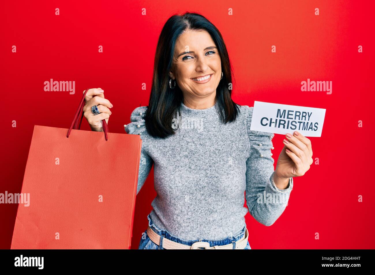 Middle age brunette woman holding merry christmas message and shopping ...