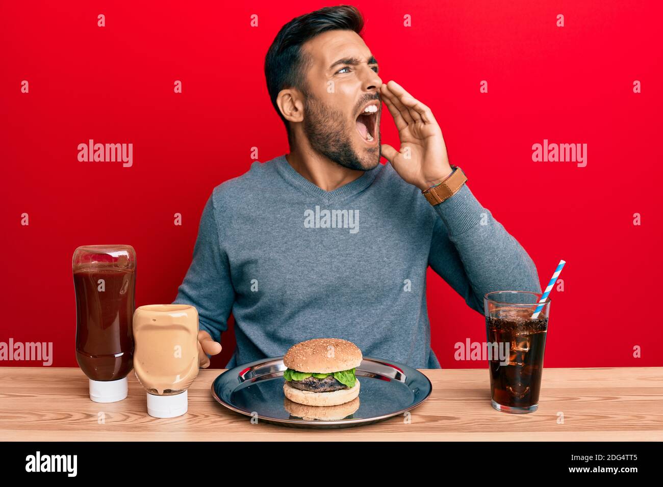 Handsome hispanic man eating a tasty classic burger and soda shouting ...
