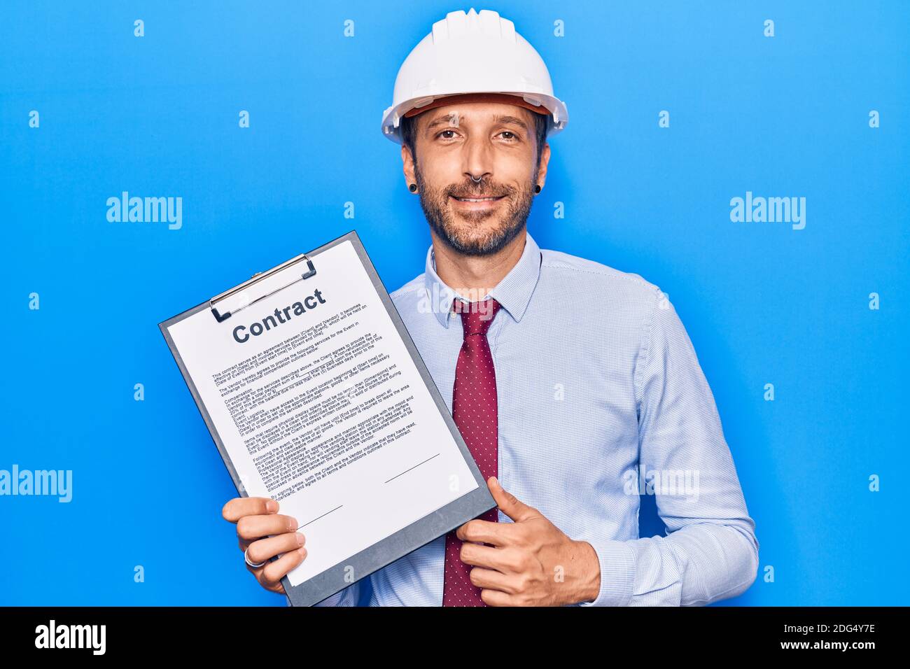 Young handsome man wearing architect hardhat holding contract document looking positive and ...