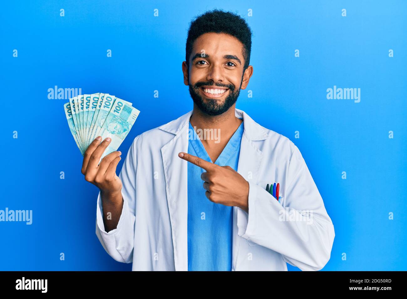 Handsome hispanic man with beard wearing doctor uniform holding 100 brazilian reals smiling ...