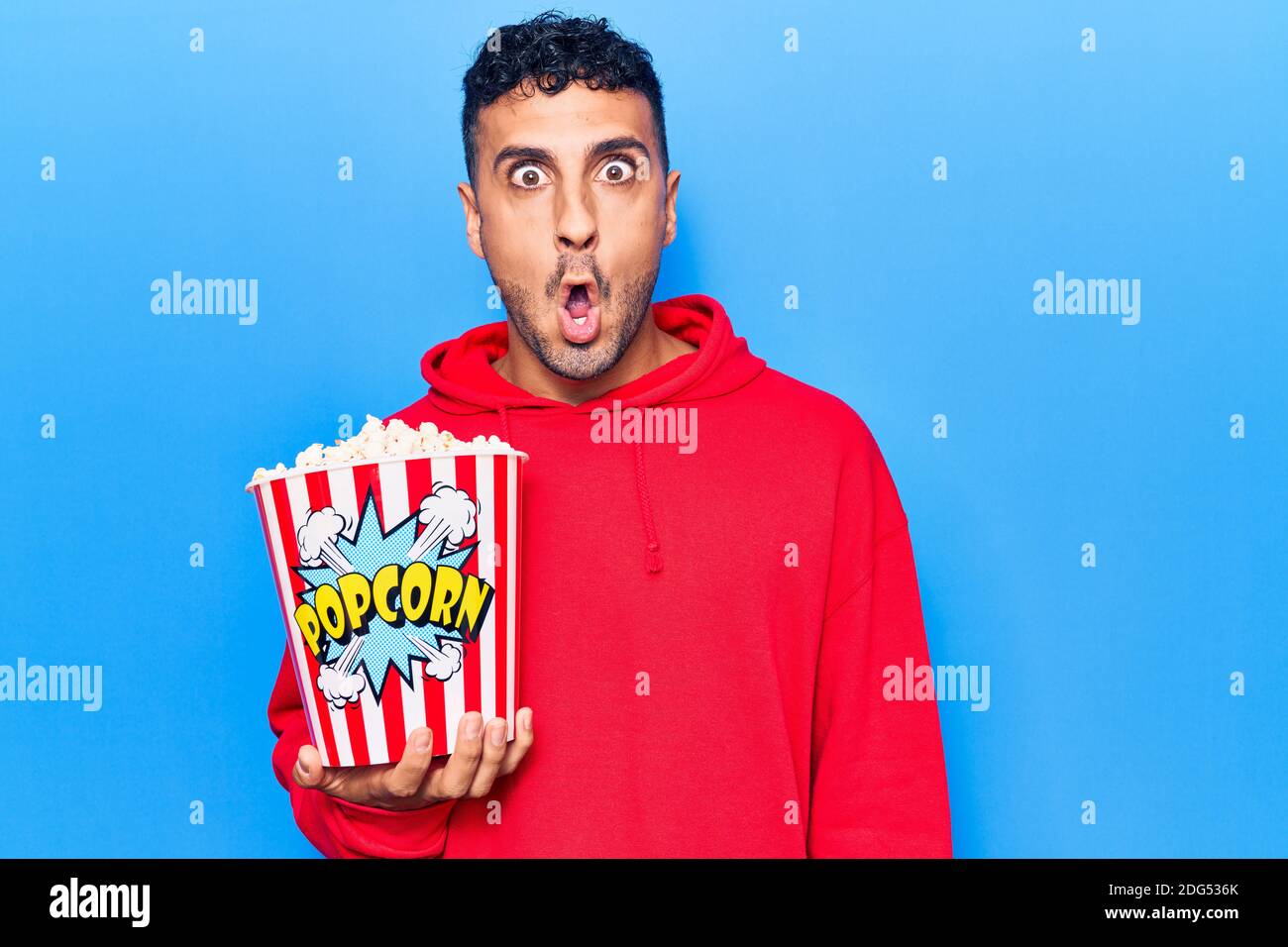 Young hispanic man holding popcorn scared and amazed with open mouth for surprise, disbelief ...