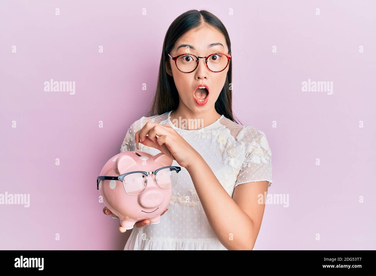 Young chinese woman holding piggy bank with glasses afraid and shocked ...
