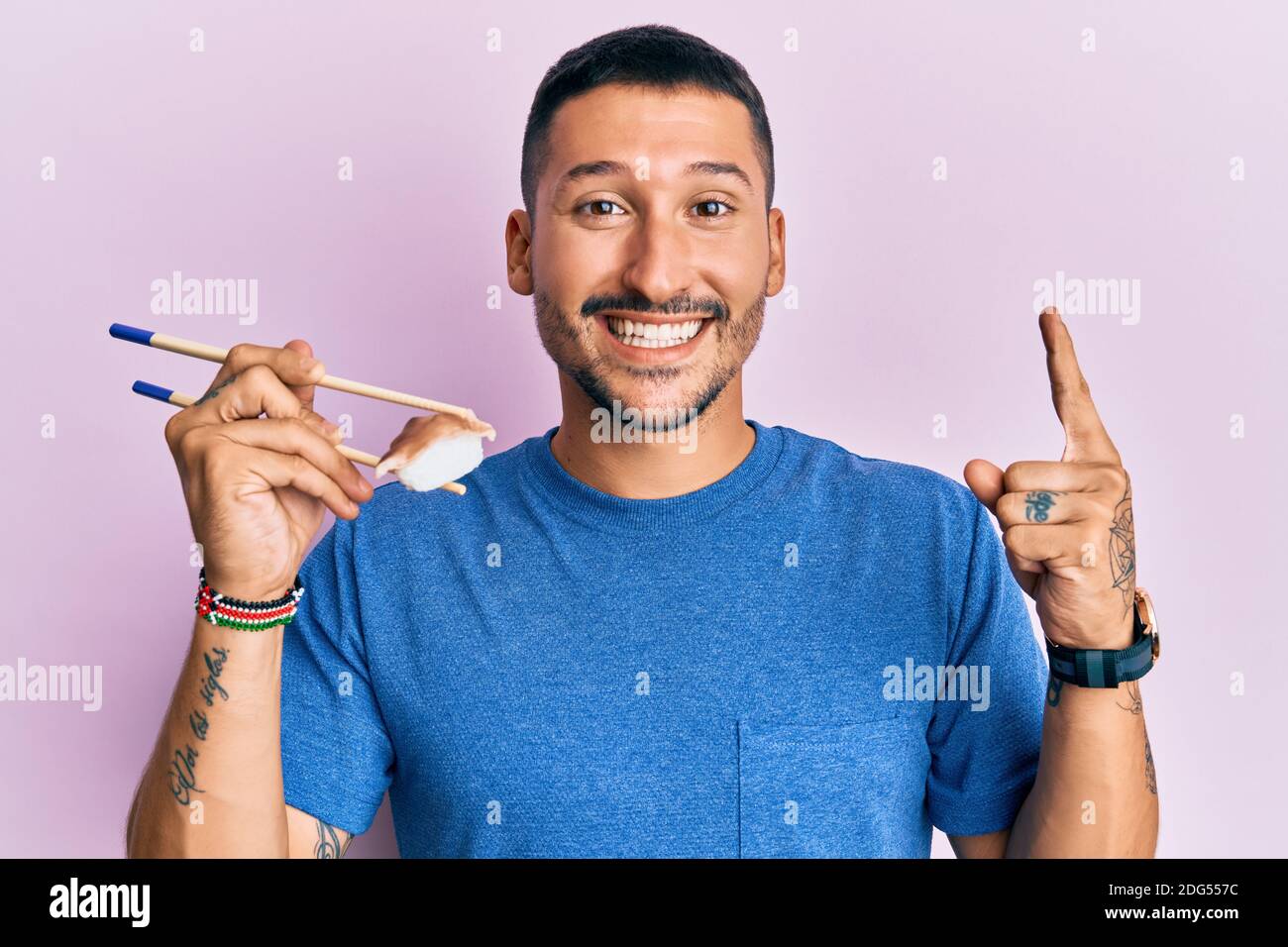 Handsome man with tattoos eating eel sushi using chopsticks smiling ...