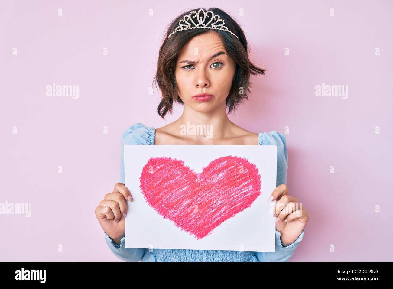Young beautiful girl wearing princess crown holding heart draw skeptic ...