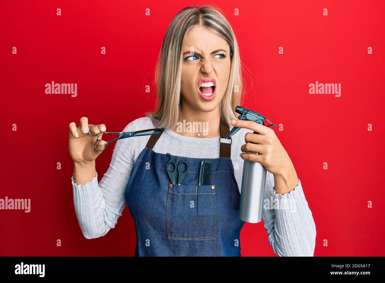 Beautiful blonde woman wearing hairdresser apron and holding scissors angry and mad screaming ...