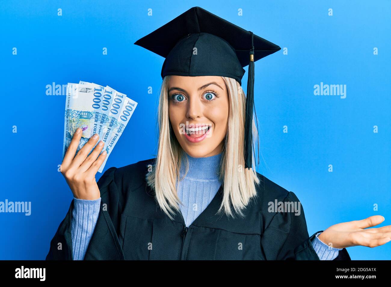 Beautiful blonde woman wearing graduation cap and ceremony robe holding ...